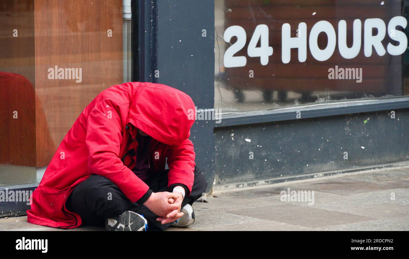 A young person sat on a street pavement curled up in a red hooded ...