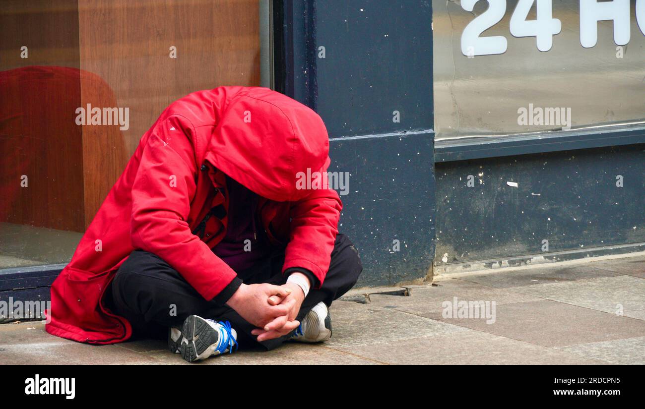 A young person sat on a street pavement curled up in a red hooded ...