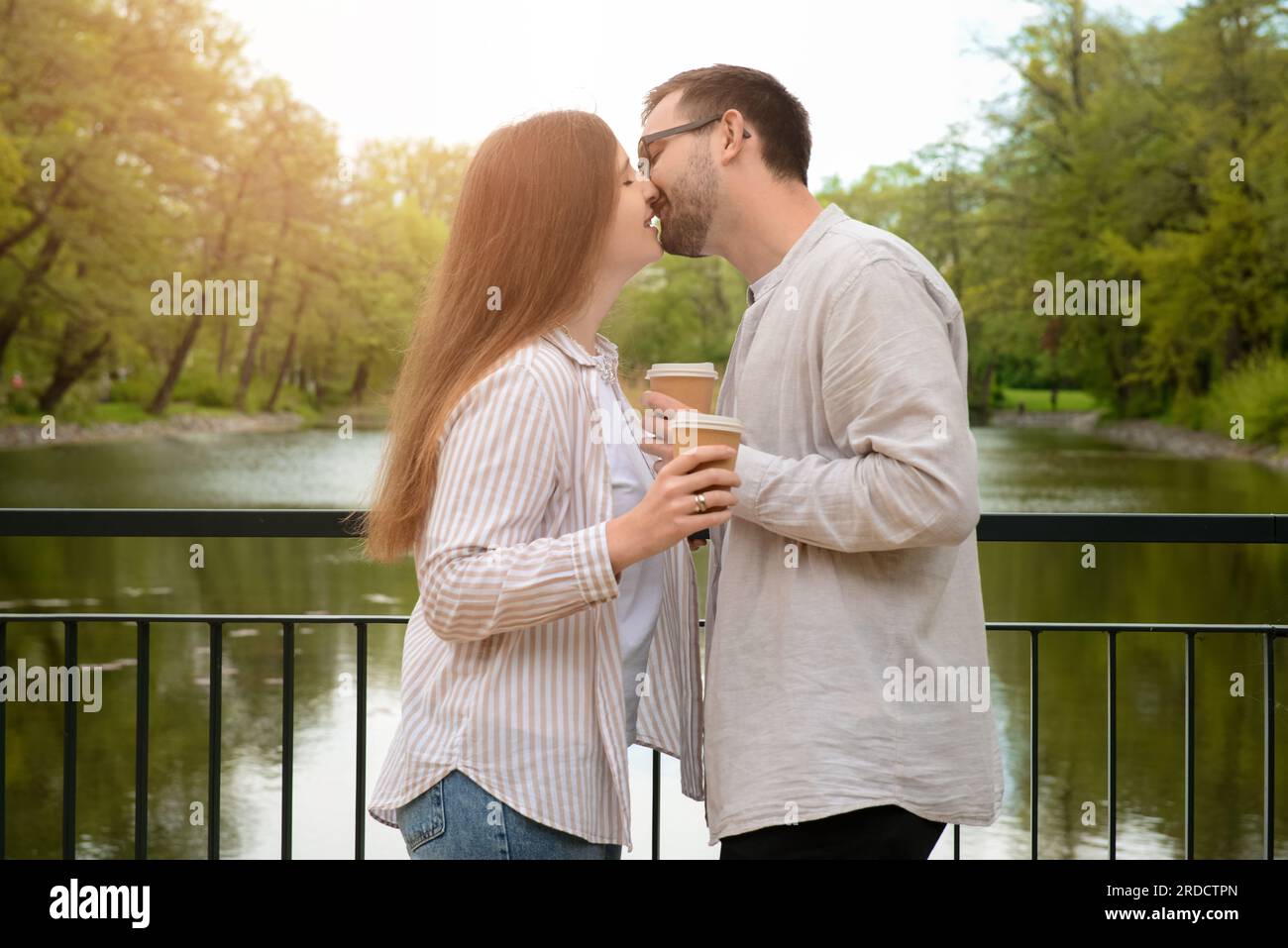 Beautiful happy couple with cups of coffee kissing on bridge in park ...