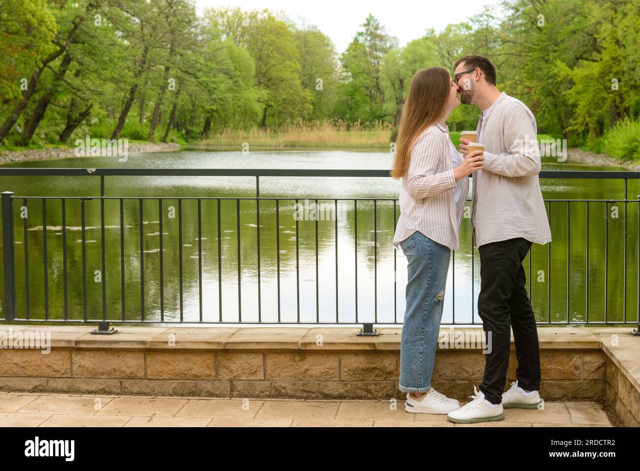 Beautiful happy couple with cups of coffee kissing on bridge in park ...