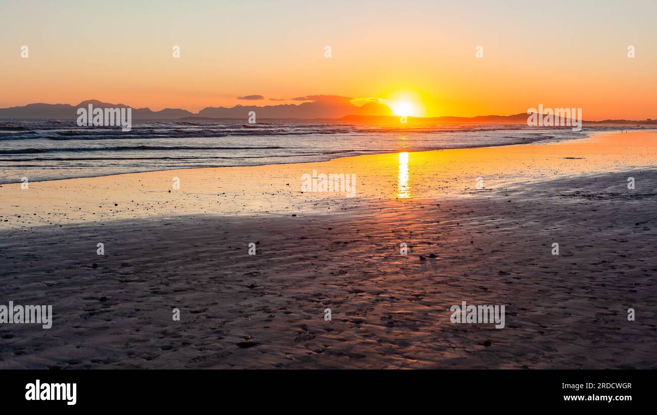 Beach ocean coastline as sun dipping water reflections behind mountains ...