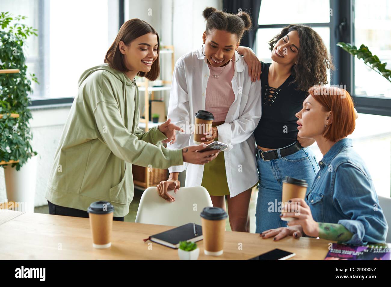 joyous young woman showing mobile phone to multicultural friends near ...