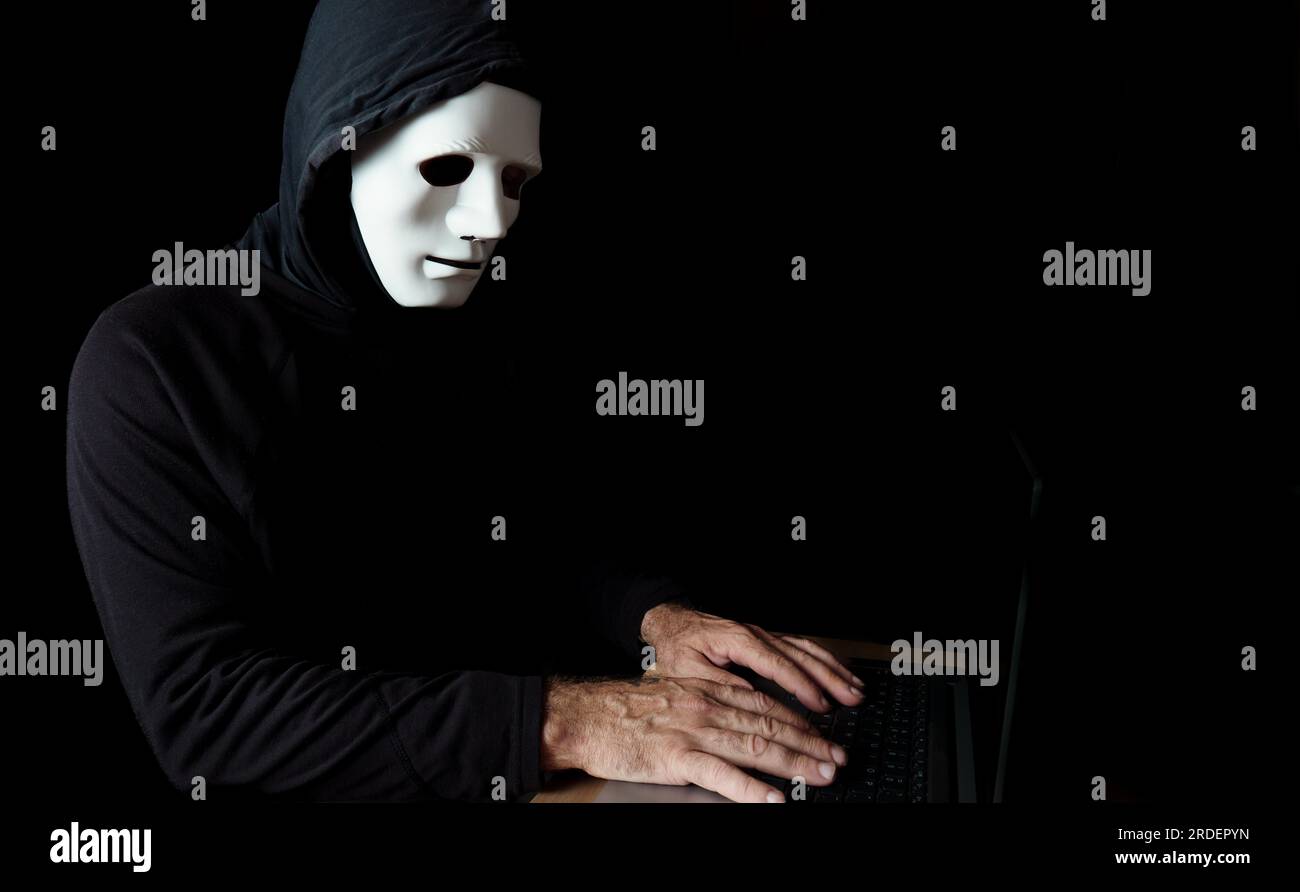 Male hacker dressed in black with a hood and a white mask using a ...