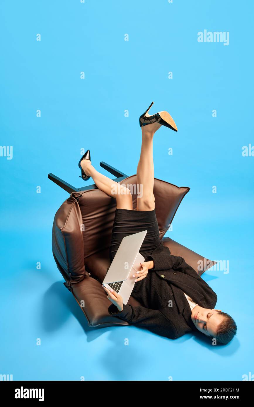 Young woman, employee lying on fallen armchair and working on laptop against blue studio ...