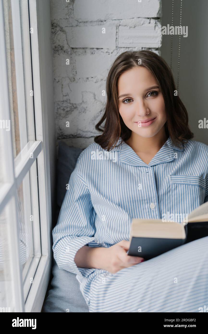 Young woman in casual clothes reading a romantic book by the window ...
