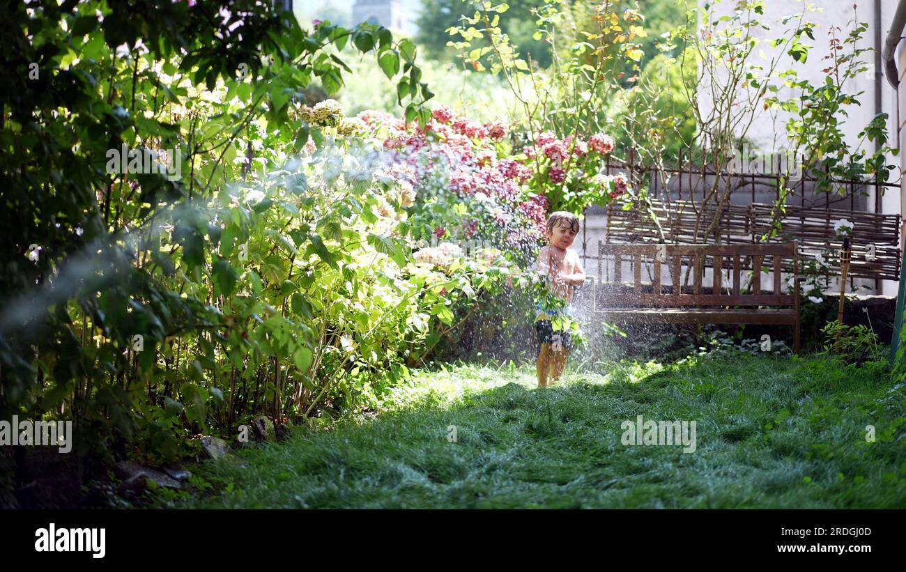 Wet little boy during hot summer day in home garden spraying water into ...