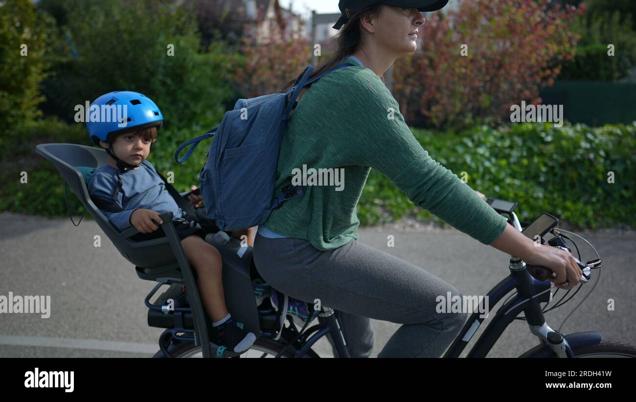 Mother riding bicycle with child in back seat outside in urban green ...