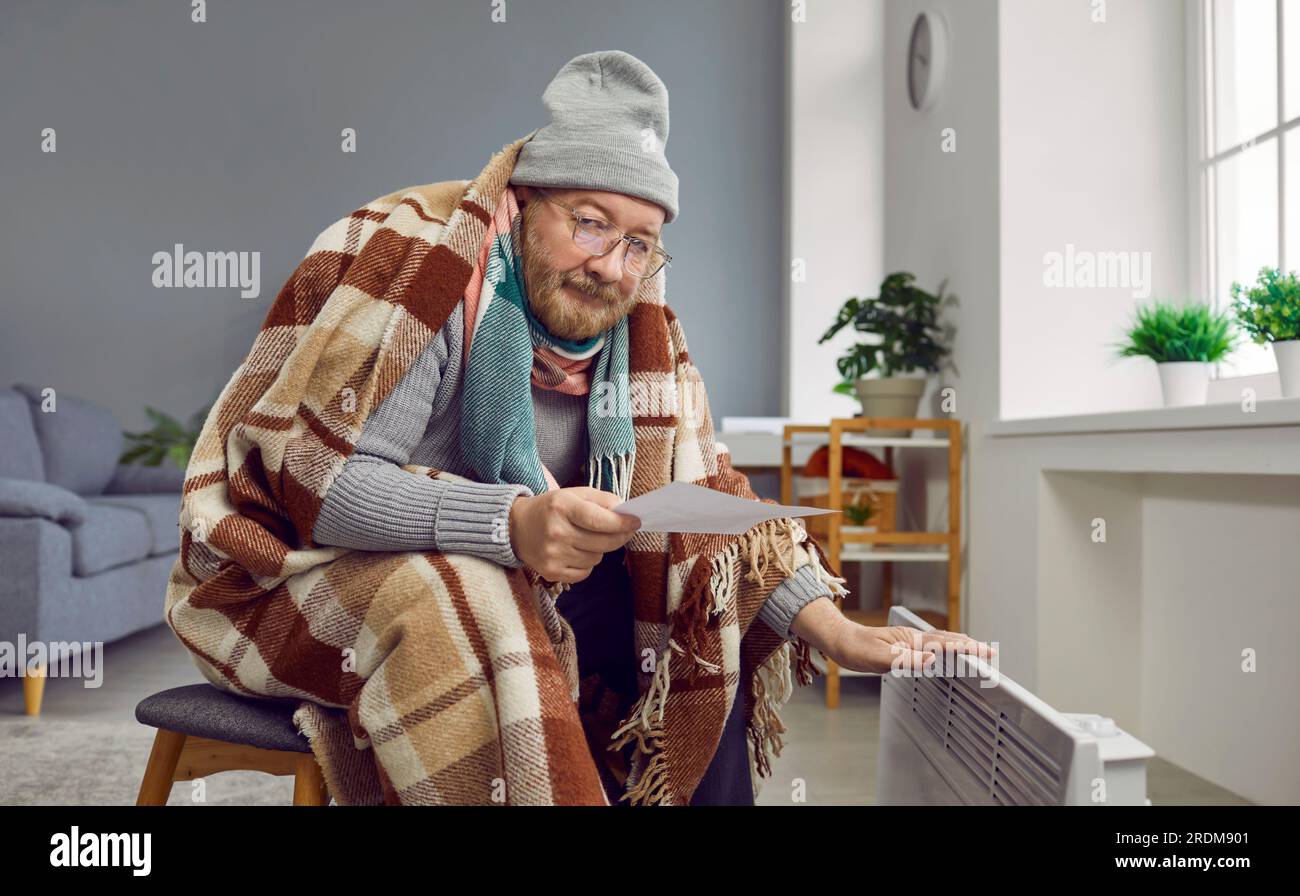Senior man sitting at home near electric heater and holding utility ...