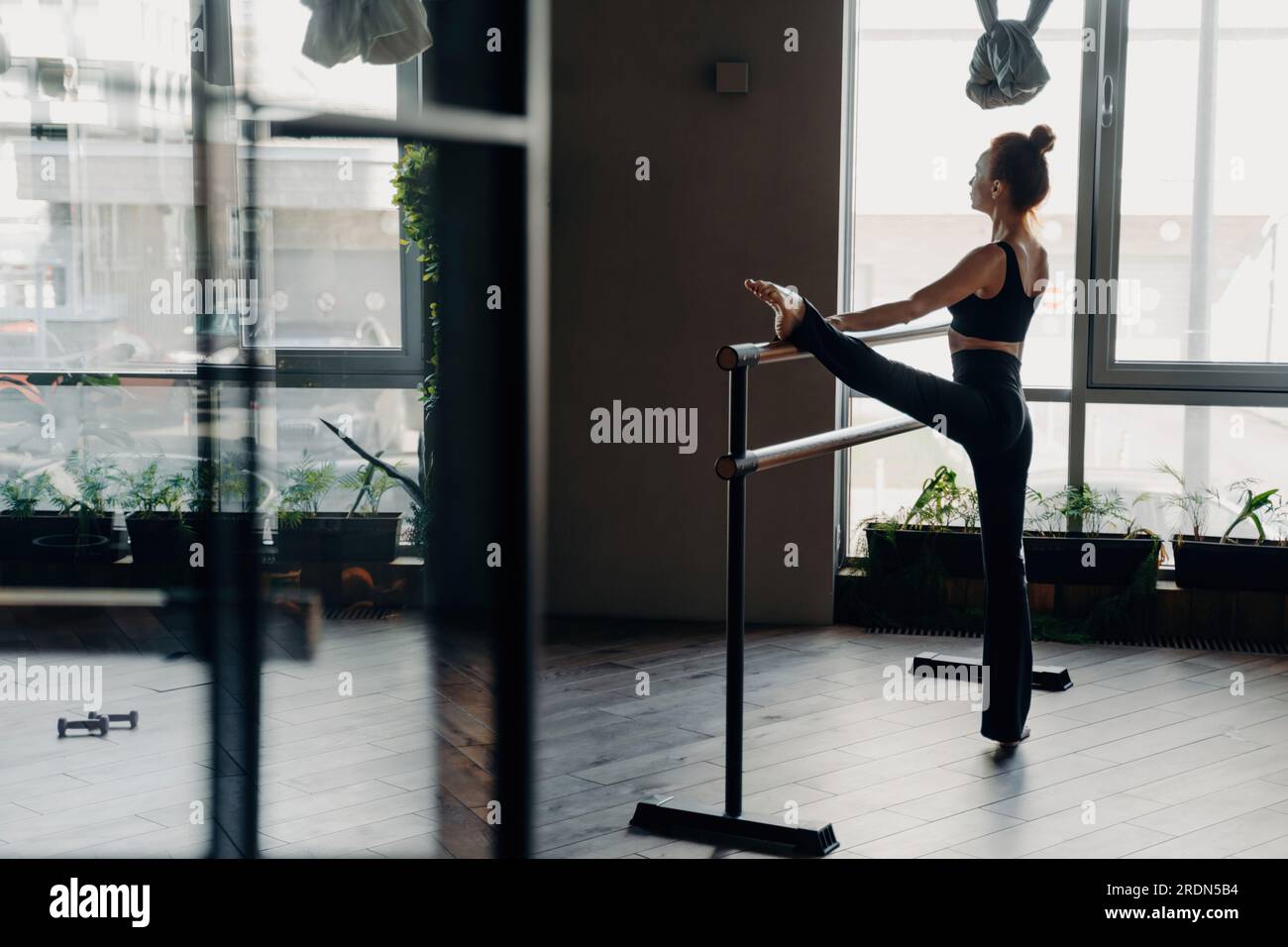 Slim red-haired ballerina stands in split position beside ballet barre ...