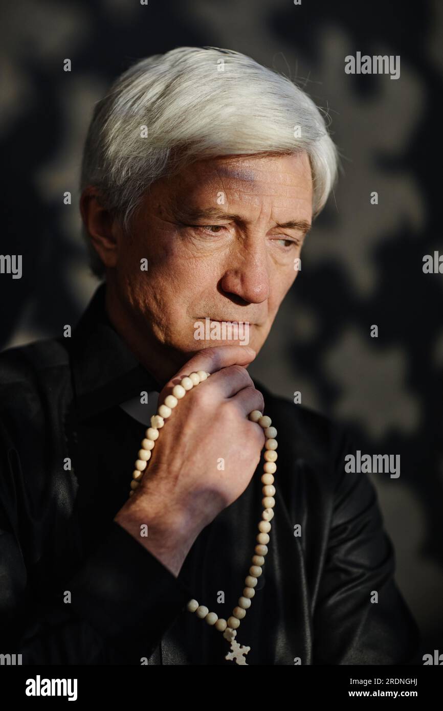 Vertical portrait of white haired senior priest holding rosary deep in ...