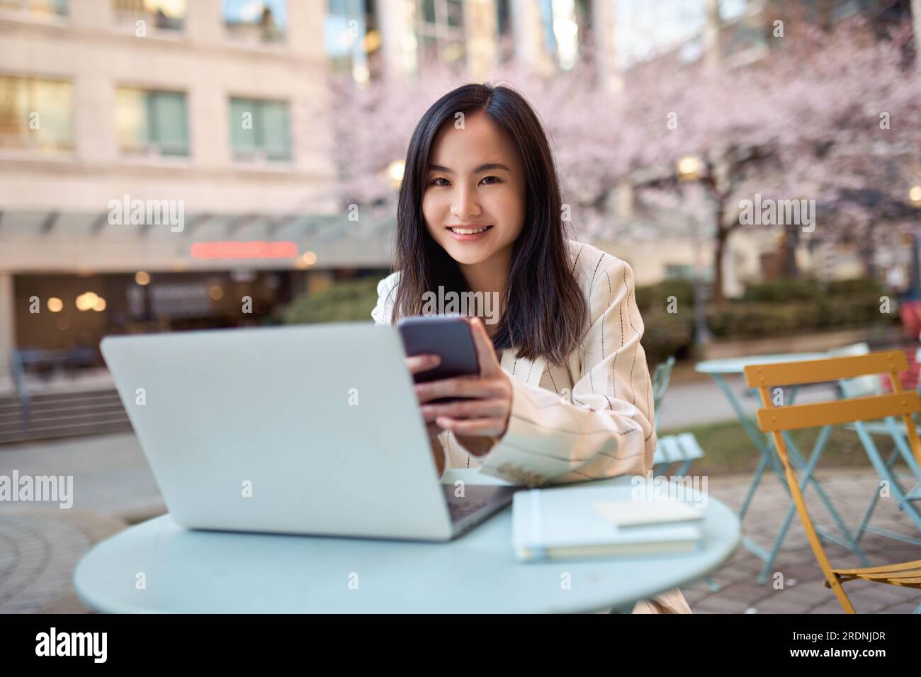 Young happy Asian woman student using mobile phone and laptop outdoors ...