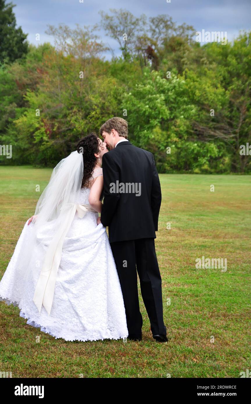 Bride and groom pause in their walk outdoors, to kiss. Both are holding ...