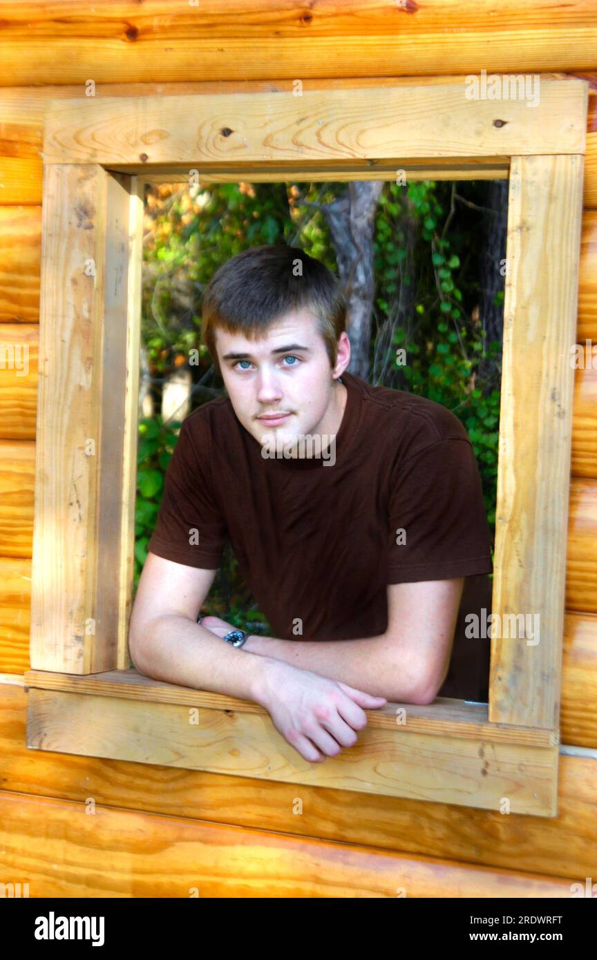 Young man leans through the window of a log cabin window. He is solemn ...