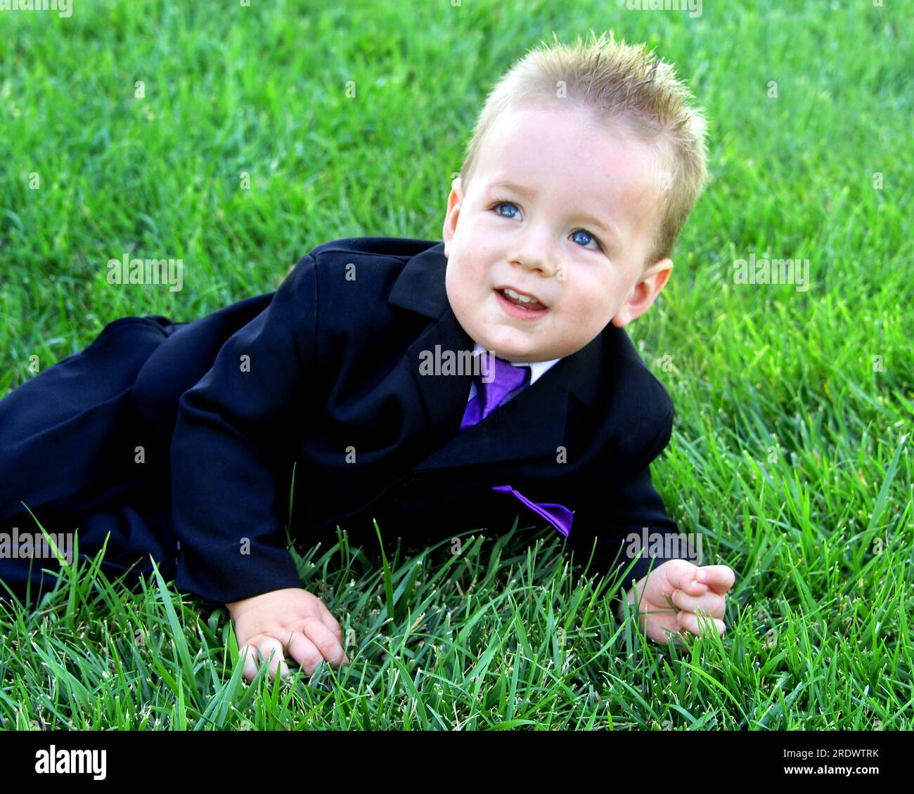 Little boy, wearing a tuxedo and tie, leans back against the grass. He ...