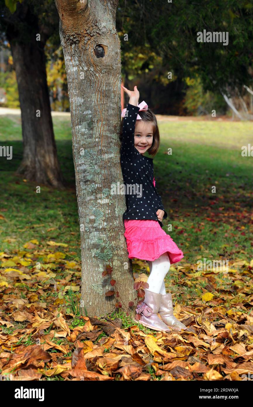 Little girl waves hello or goodbye as she stands besides a tree ...