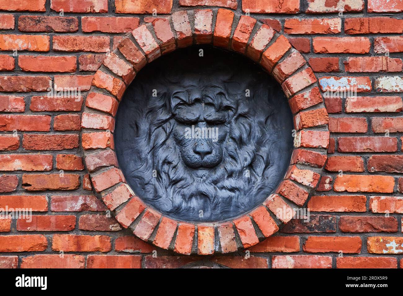 Faded red brick wall with circular opening to lion head with mane motif ...