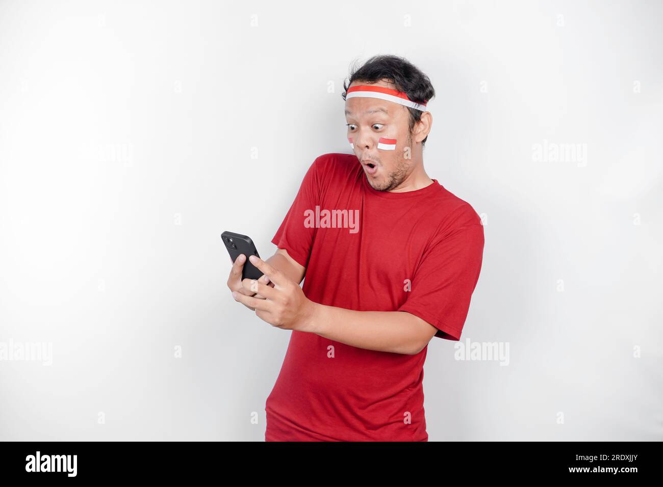A portrait of a shocked Asian man wearing headband, holding his phone ...