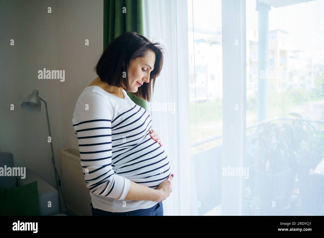 Smiling pregnant woman touching stomach and standing by window Stock ...