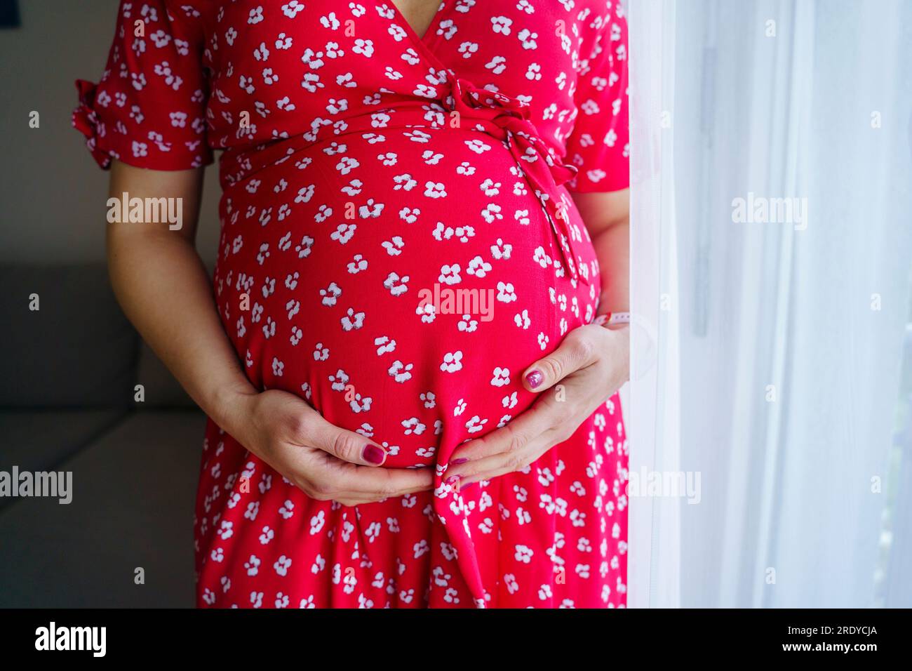 Pregnant woman touching stomach standing in living room by window at ...