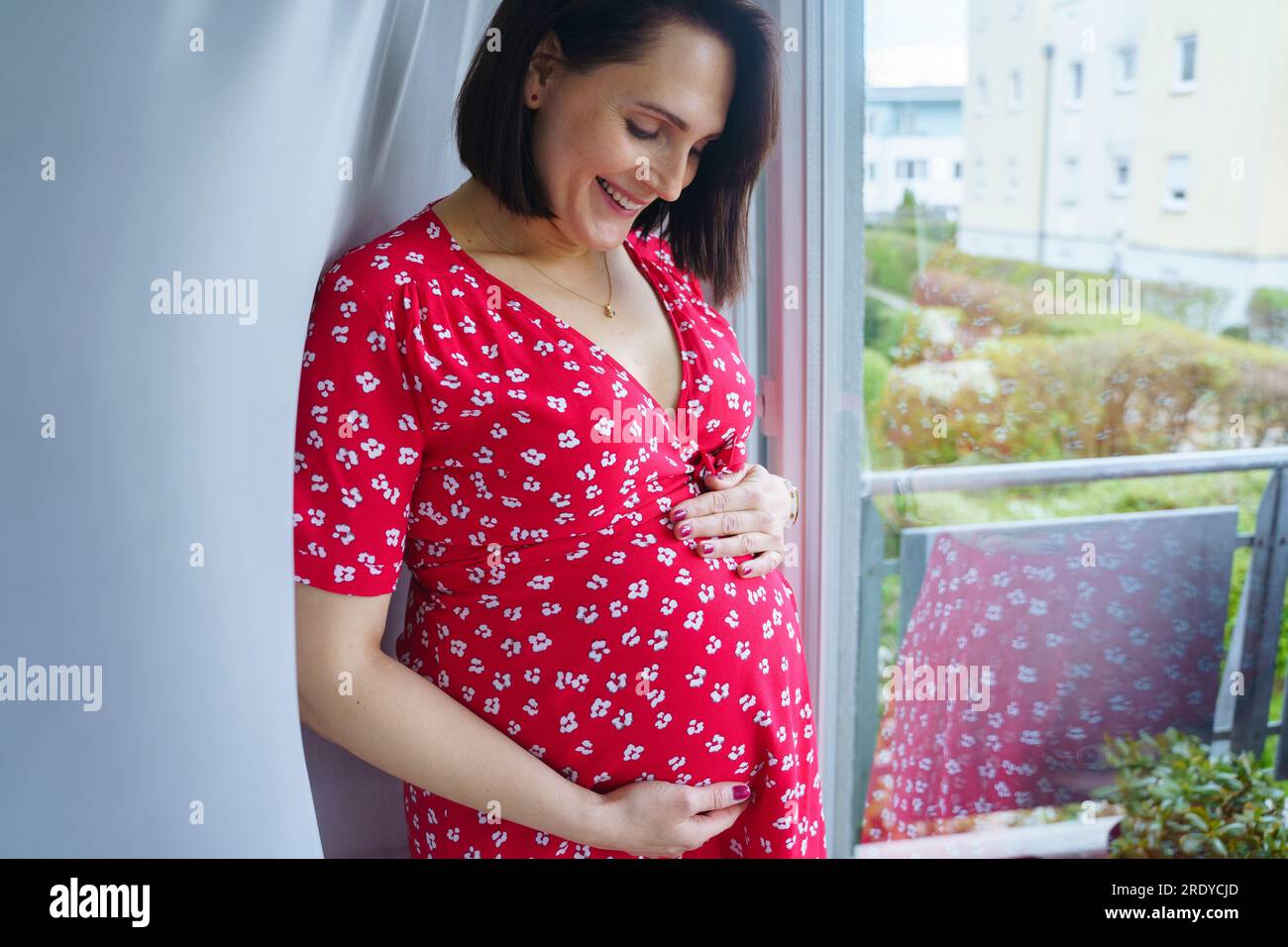 Happy pregnant woman touching stomach and standing by window at home ...