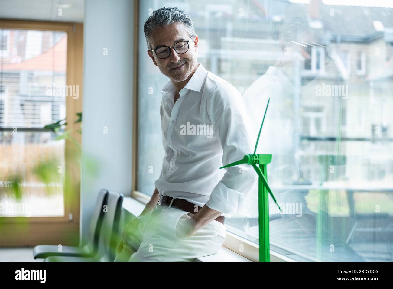 Smiling businessman looking at wind turbine model leaning on window ...