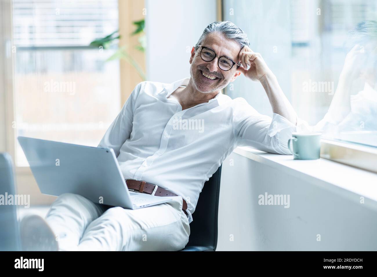 Happy businessman with laptop leaning head on window sill at office ...
