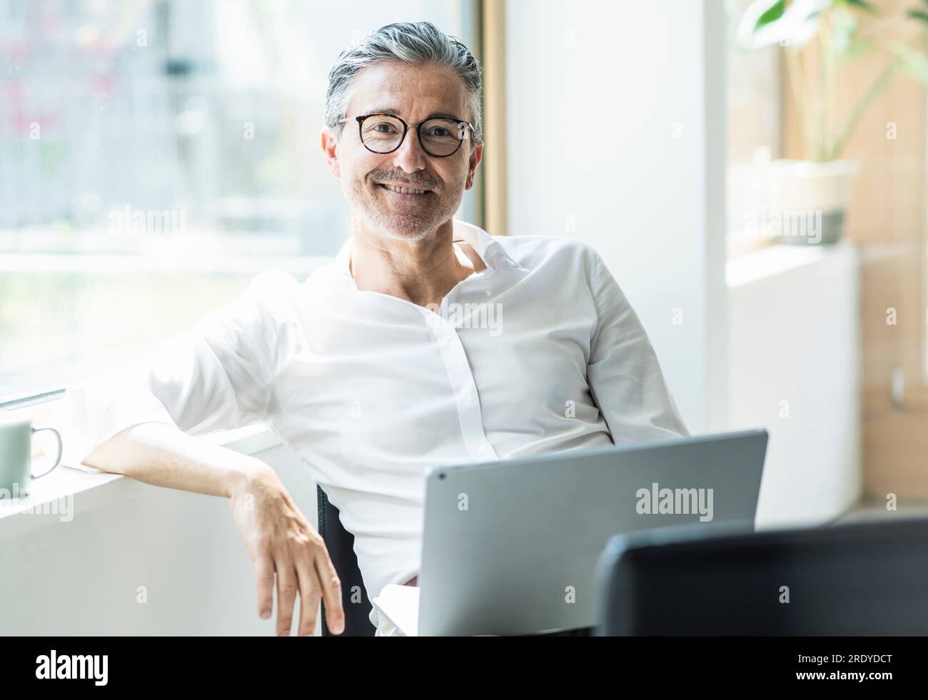 Smiling businessman sitting with laptop leaning head on window sill at ...