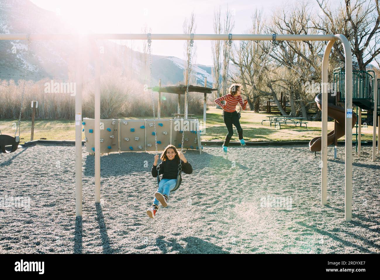 Two teen girls swing on swings at playground with snow in background ...