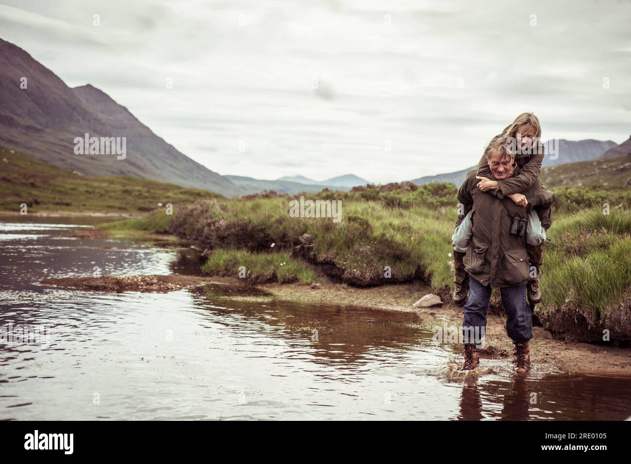 retired couple piggy back carry across remote mountain fishing river ...
