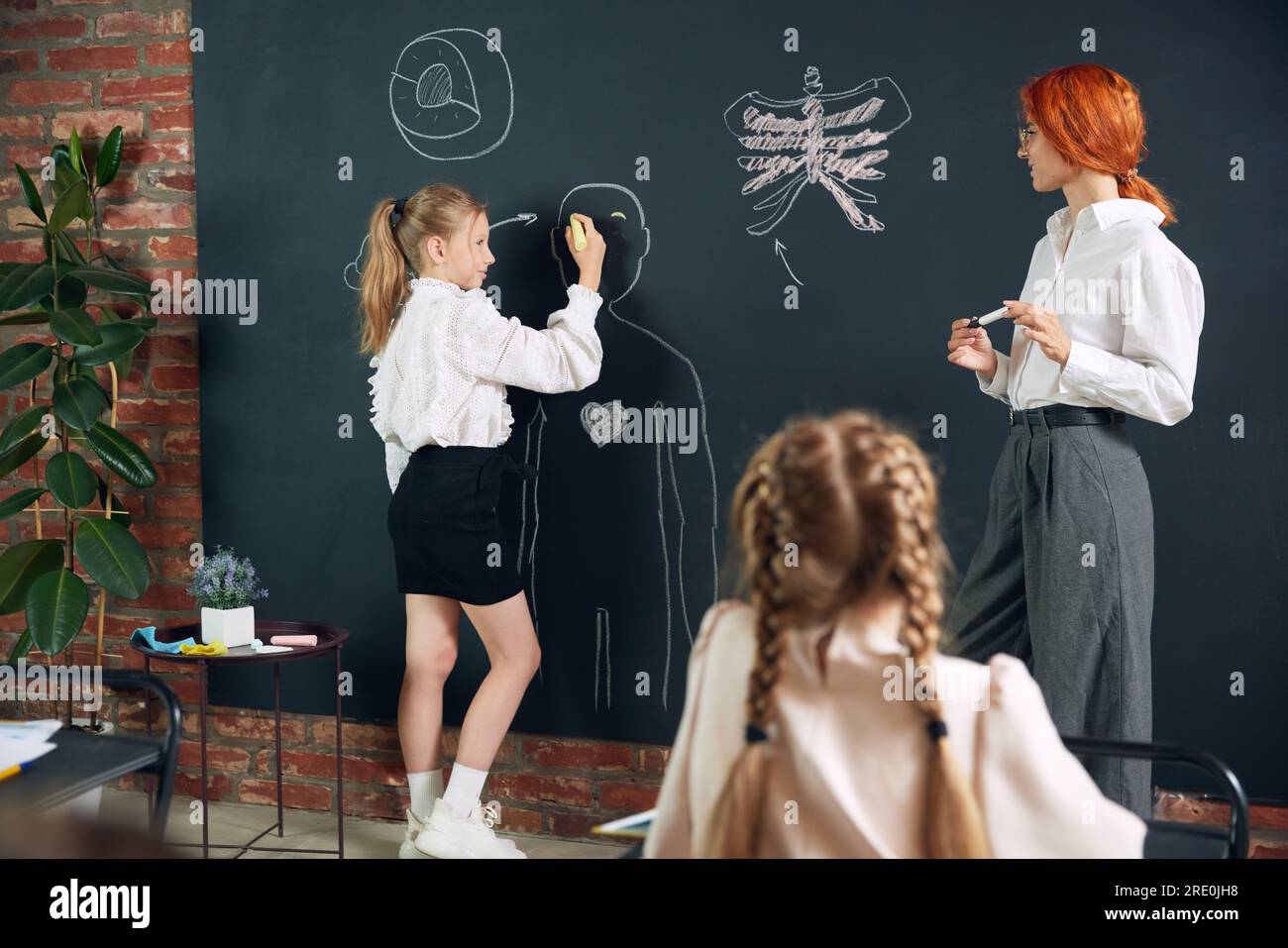 Girl, student of primary school standing at chalkboard with teacher and ...