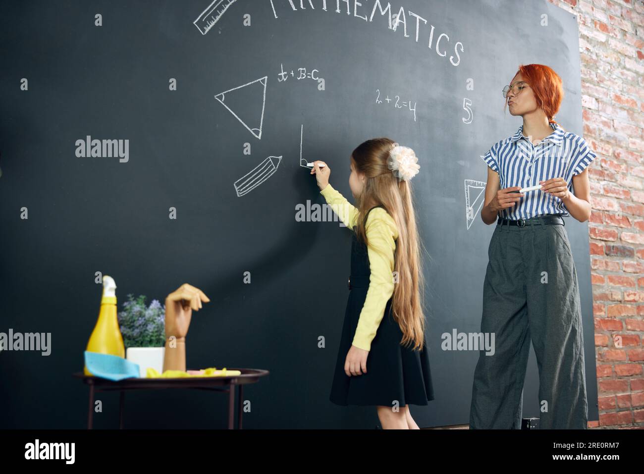 Little smart girl standing by chalkboard with teacher, writing and ...