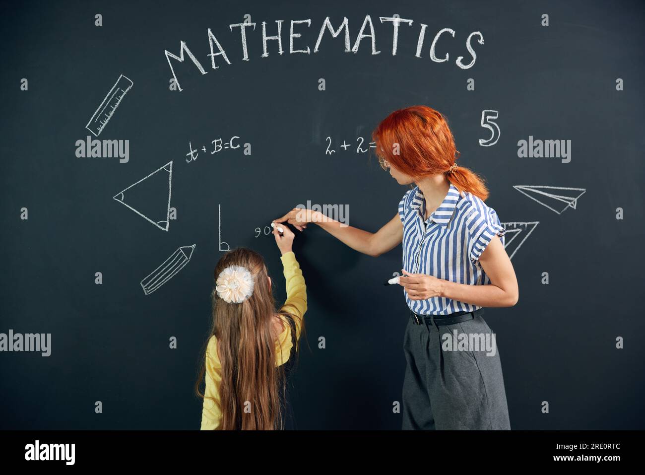 Little smart girl standing by chalkboard with teacher, writing and ...
