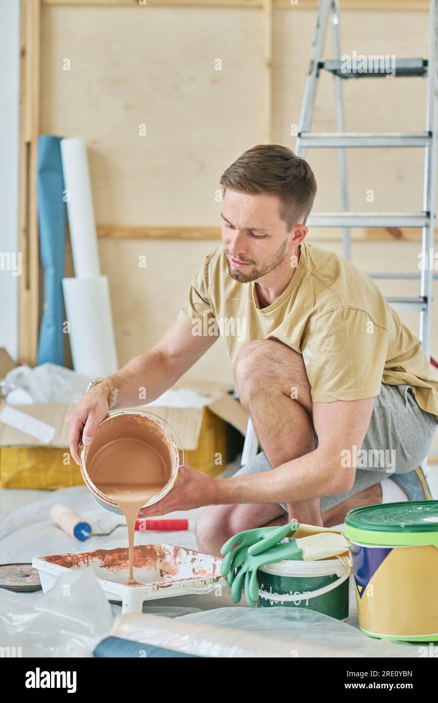 Young man pouring liquid beige paint into plastic tray during ...