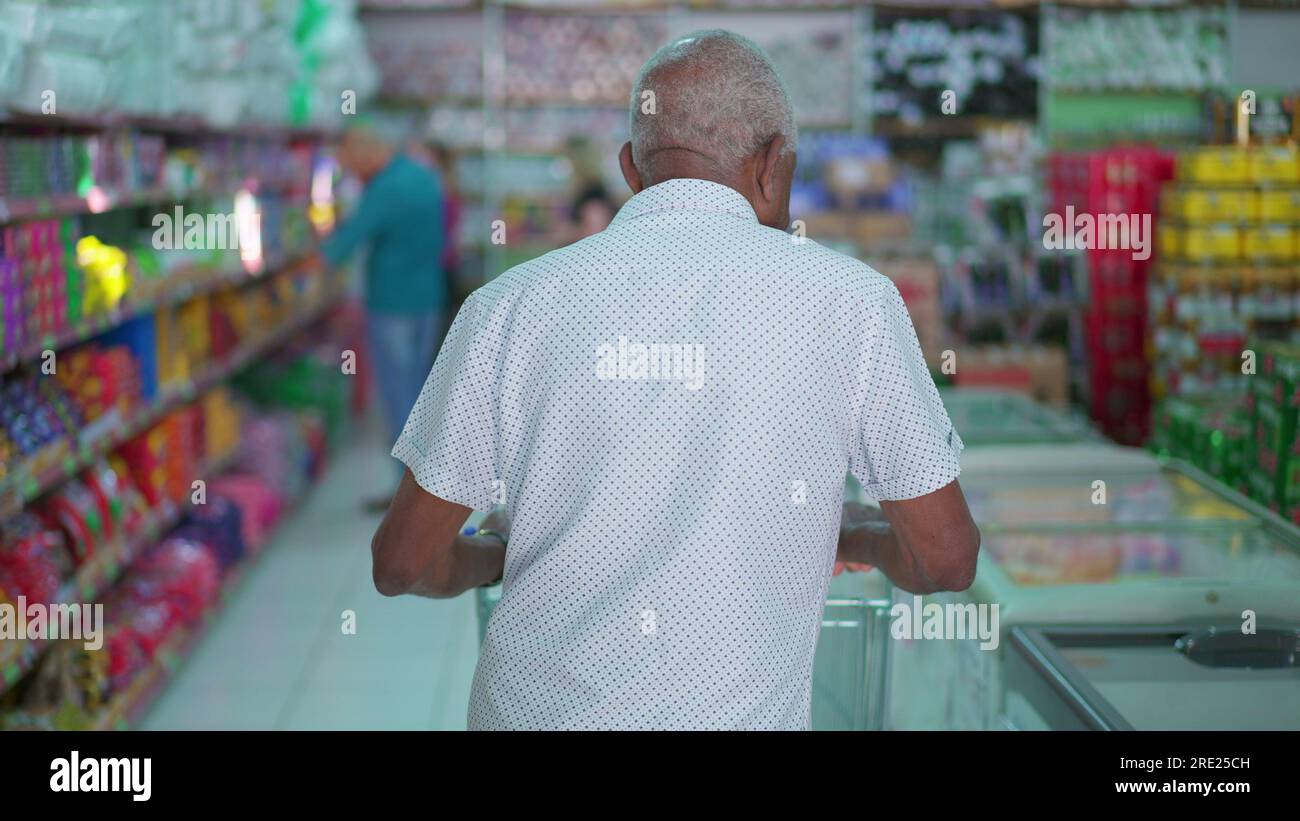 Back of senior black shopper browsing products while walking through grocery store pushing ...