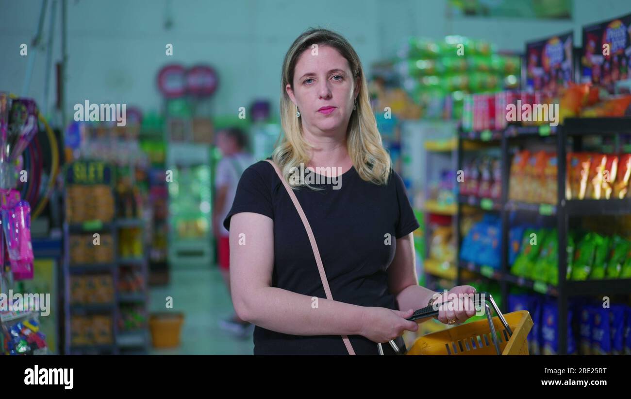 Female consumer standing in supermarket holding basket with serious ...
