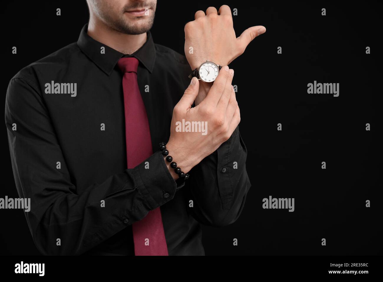 Elegant young man looking at wristwatch on black background, closeup ...