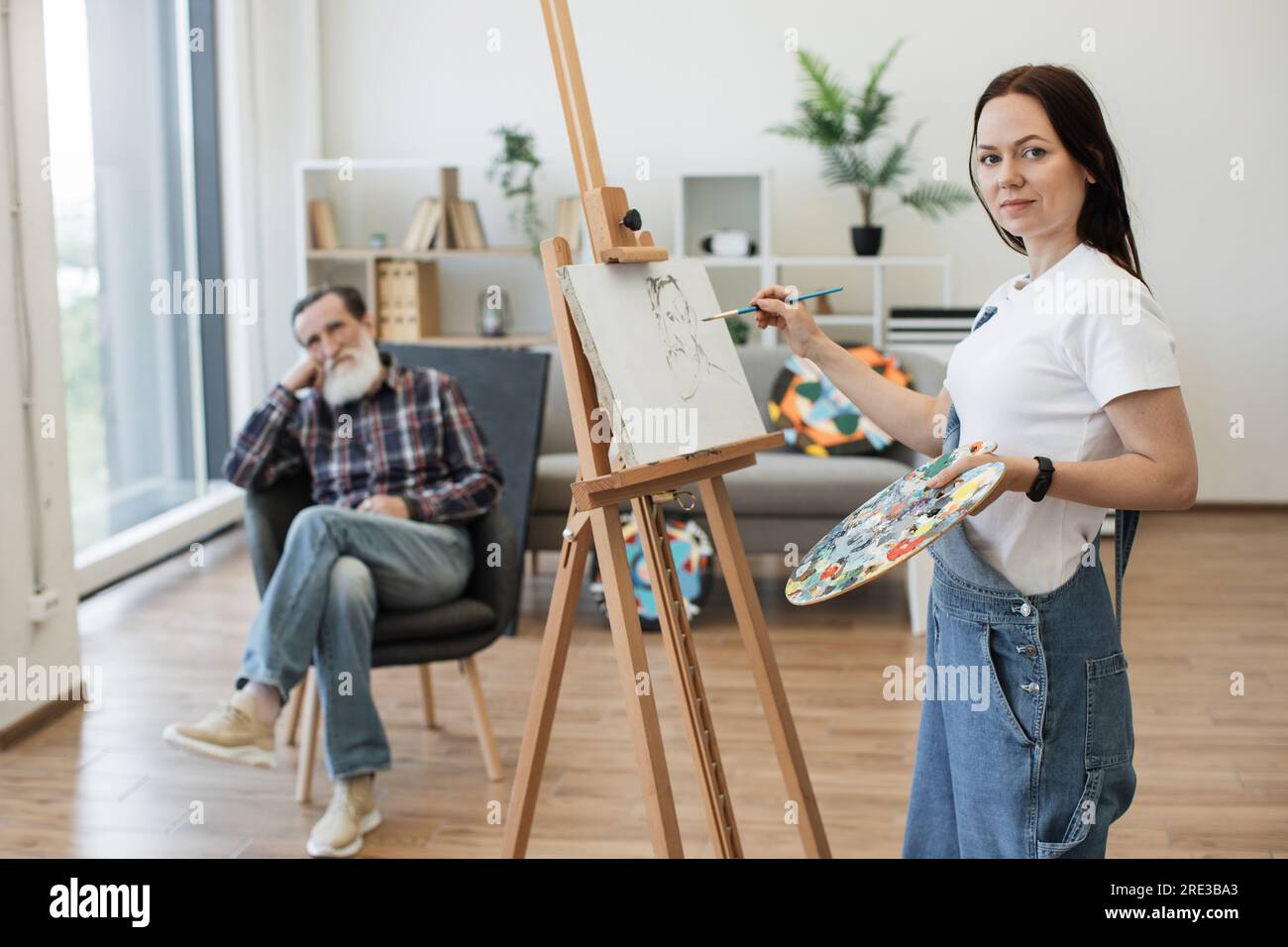 Self-employed female painter in denim overalls depicting sitter head ...