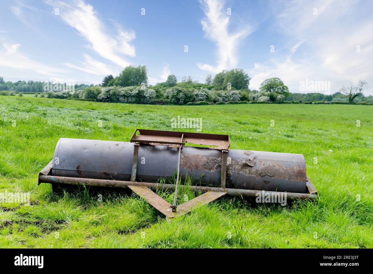 An old rusty farming roller lying in a field with a copse of bushes and ...