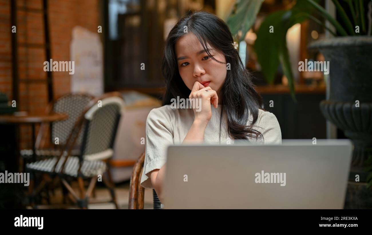 A doubtful and thoughtful young Asian woman is looking at her laptop ...