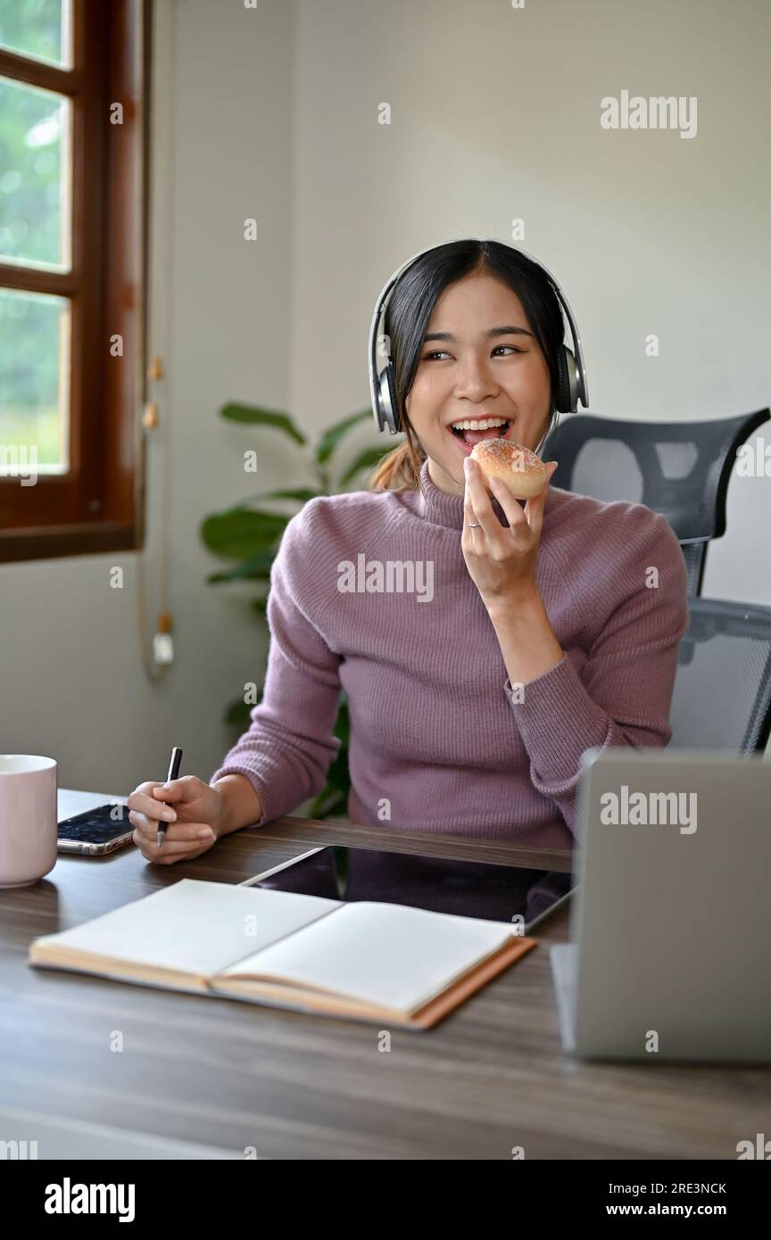 Beautiful young Asian girl eating a donut while studying at her study ...