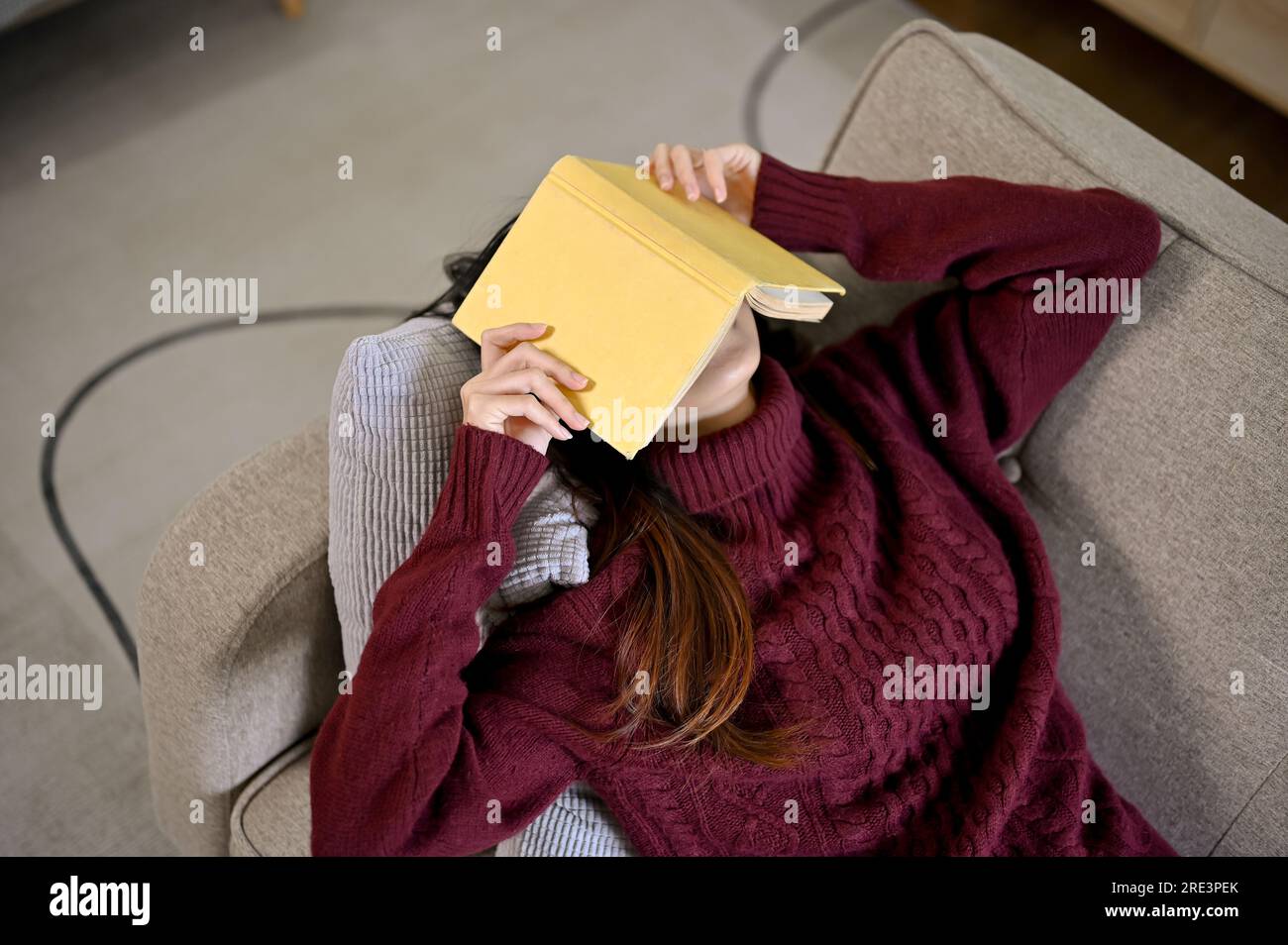 Young Asian woman laying down on the sofa with book cover on her face, taking a nap, relaxing in ...