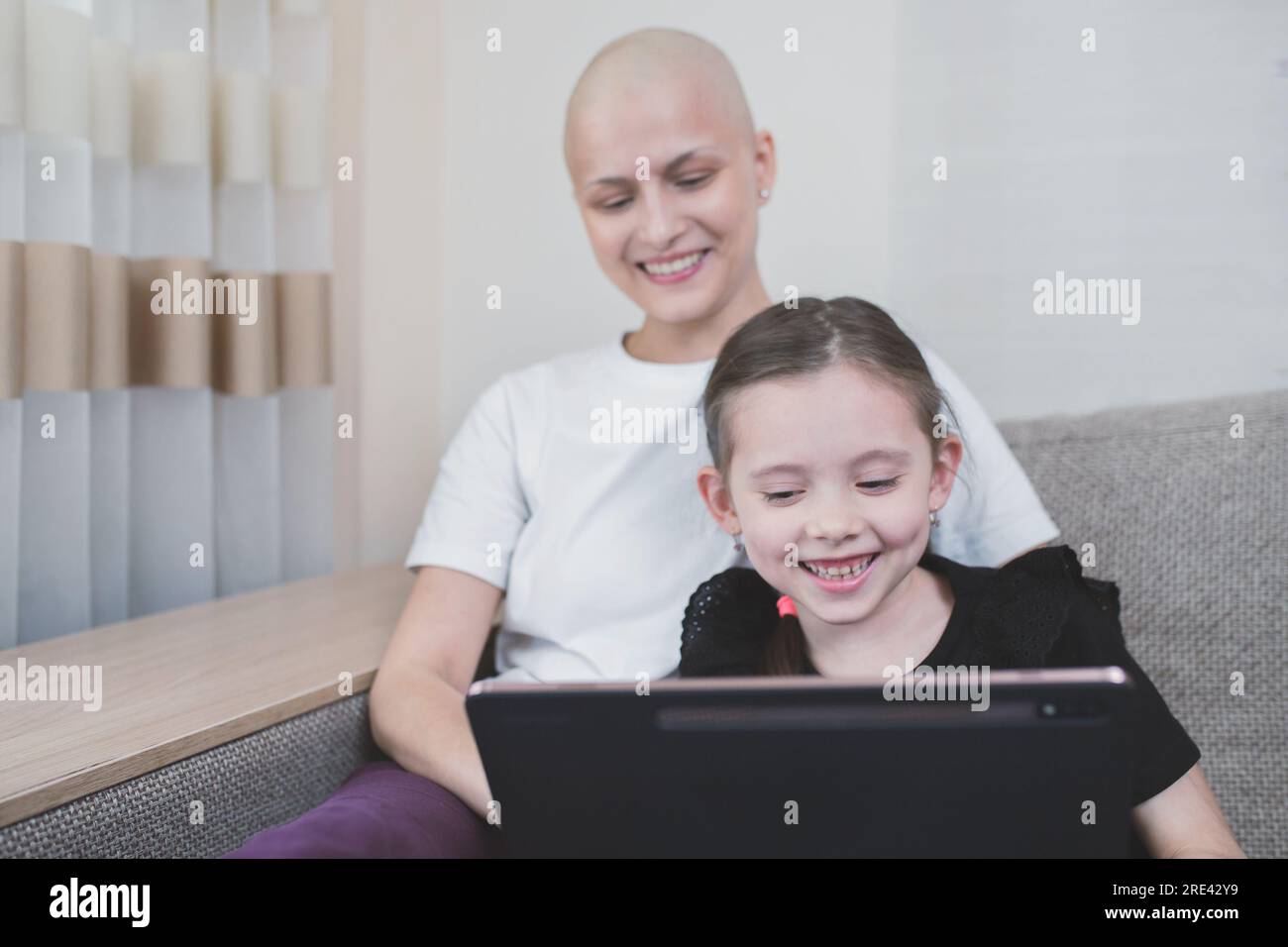 Happy bald woman with alopecia smiles and joyfully spends time with her child watching cartoons ...