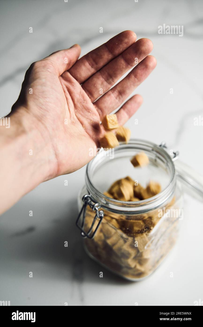Brown sugar cubes in a man's hand and in the jar on white marble ...