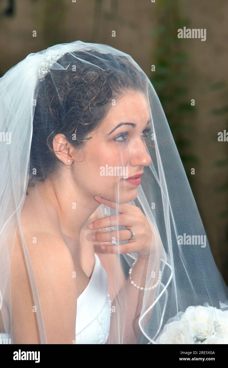 Beautiful bride sits outdoors with veil over her face. She is holding a ...