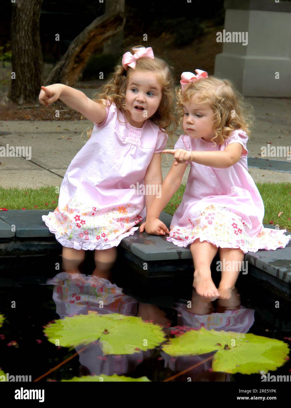 Two beautiful girls sit side by side on a lilly pad pool. They are ...