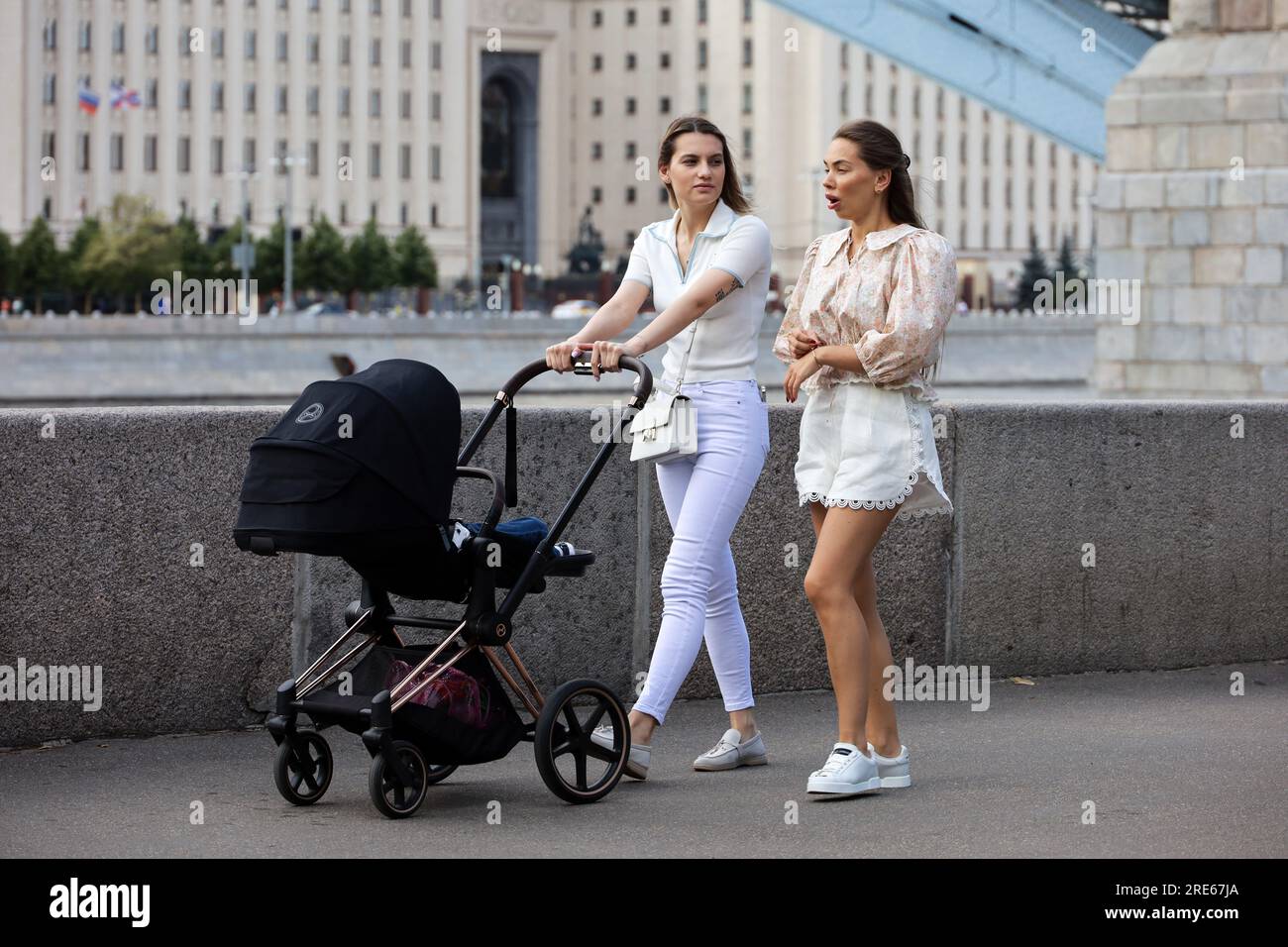 Two young women walking with baby pram in summer city on embankment of ...