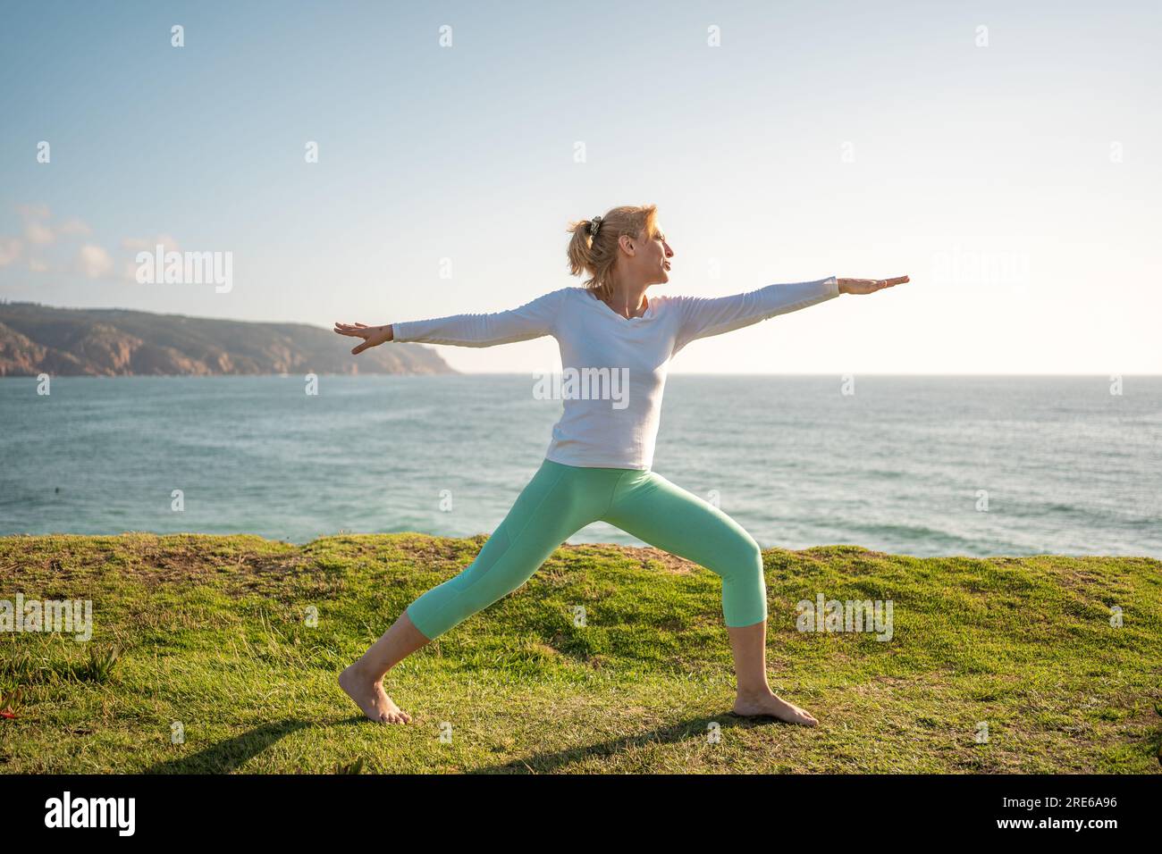 Senior woman pensioner practicing yoga warrior pose on beach. Performing virabhadrasana enjoying ...