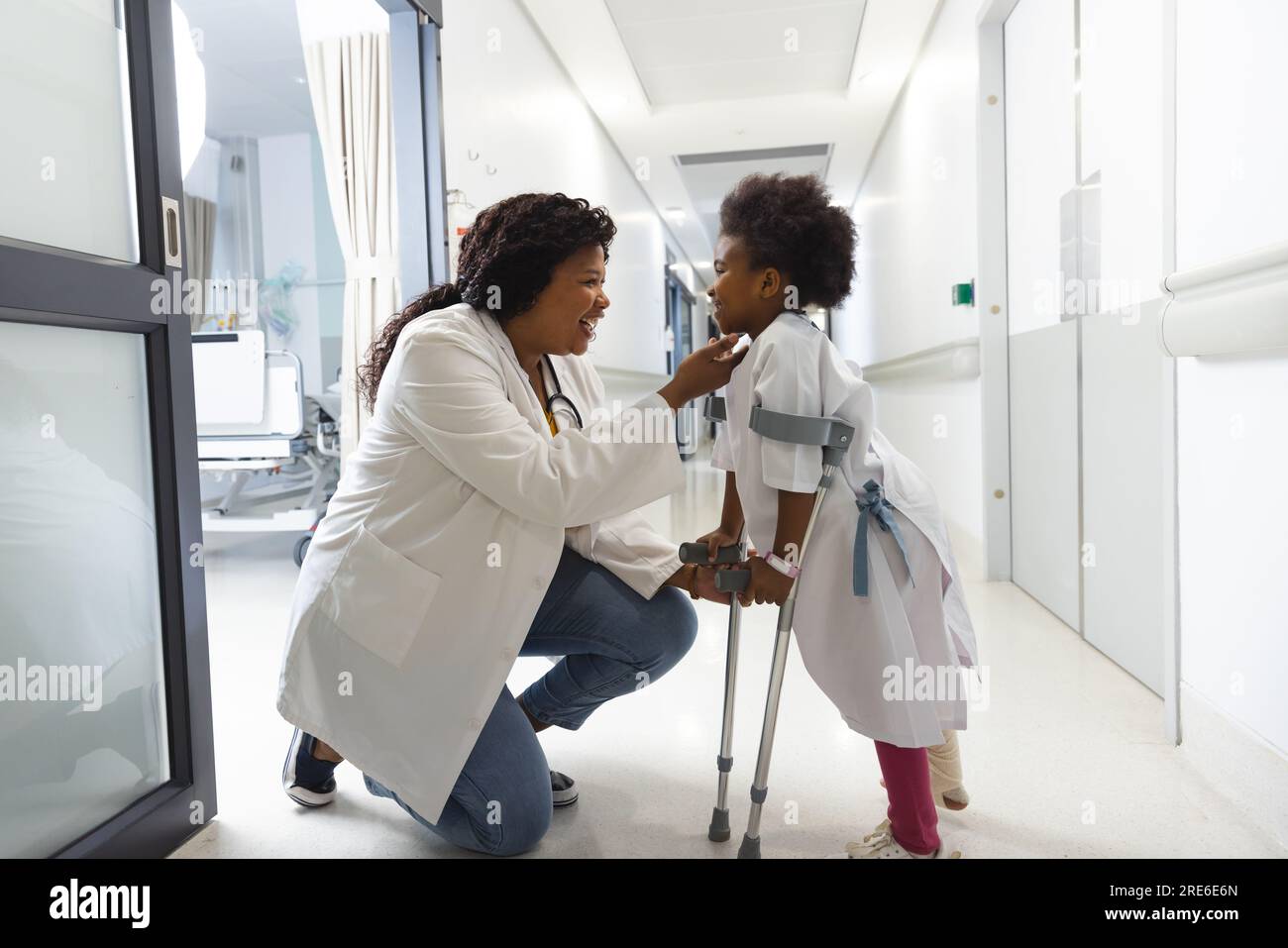 African american female doctor and girl patient walking with crutches in corridor at hospital ...