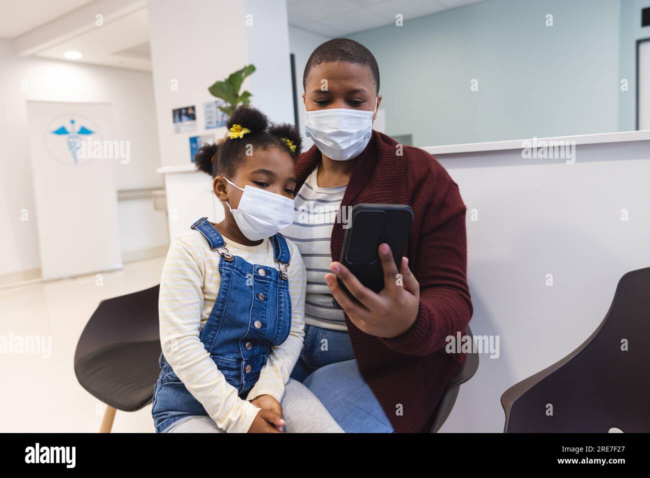 African american mother and daughter wearing face masks using smartphone in waiting room at ...