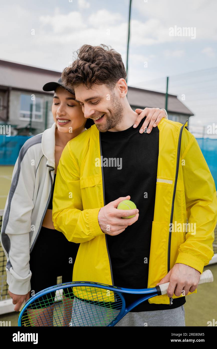 happy woman in cap and active wear hugging boyfriend holding racket and ...